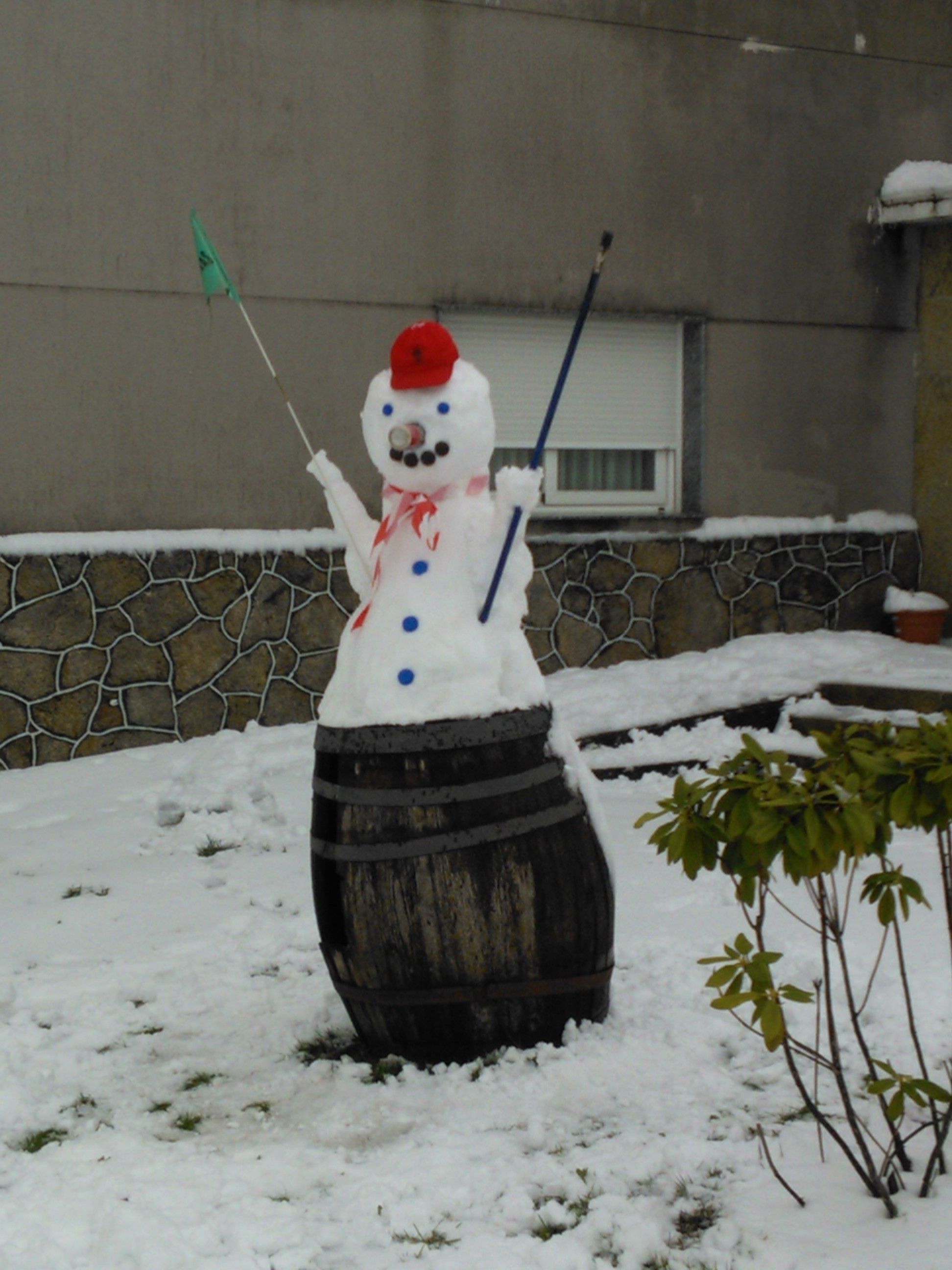Escultura de boneco de neve em área externa coberta de neve, com uma parede de pedra e uma janela ao fundo.