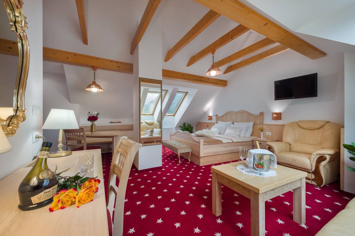 Quarto com cama, sofá, mesa com garrafa e flores, televisão e tapete vermelho com padrão de estrelas.