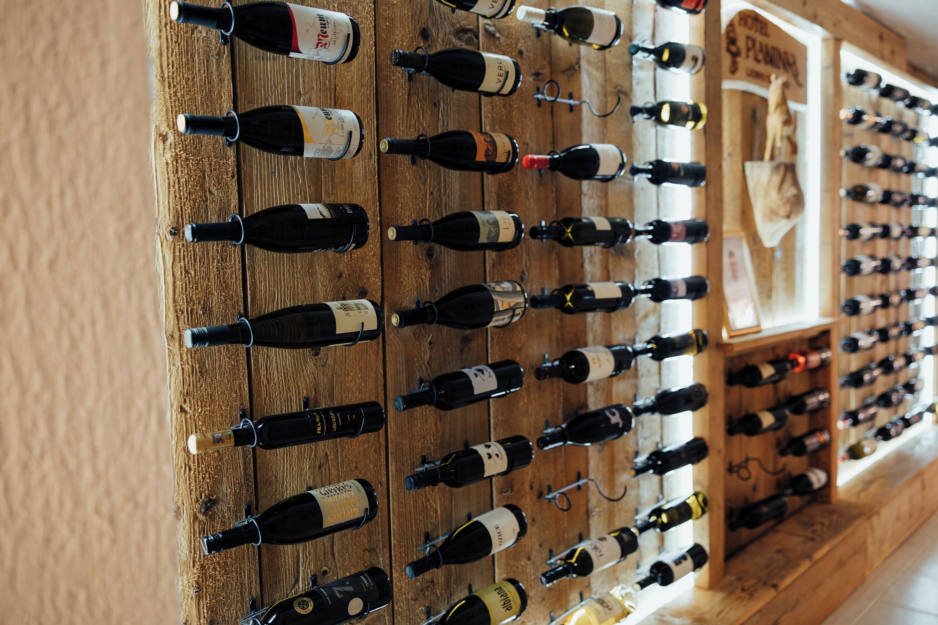 Uma parede de garrafas de vinho dispostas em fileiras, com uma prateleira de madeira e uma placa ao fundo.