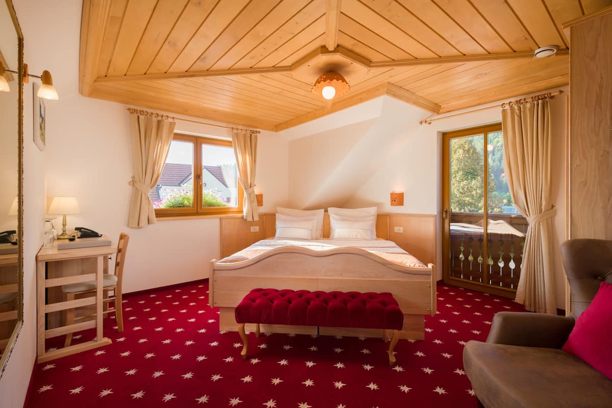 Quarto com teto de madeira, cama com roupa de cama branca, tapete vermelho, um banco, uma cadeira e uma escrivaninha com abajur.