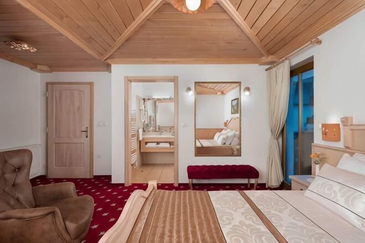 Quarto com teto de madeira, cama com roupa de cama estampada, tapete vermelho, um banco de madeira, um espelho e uma porta.
