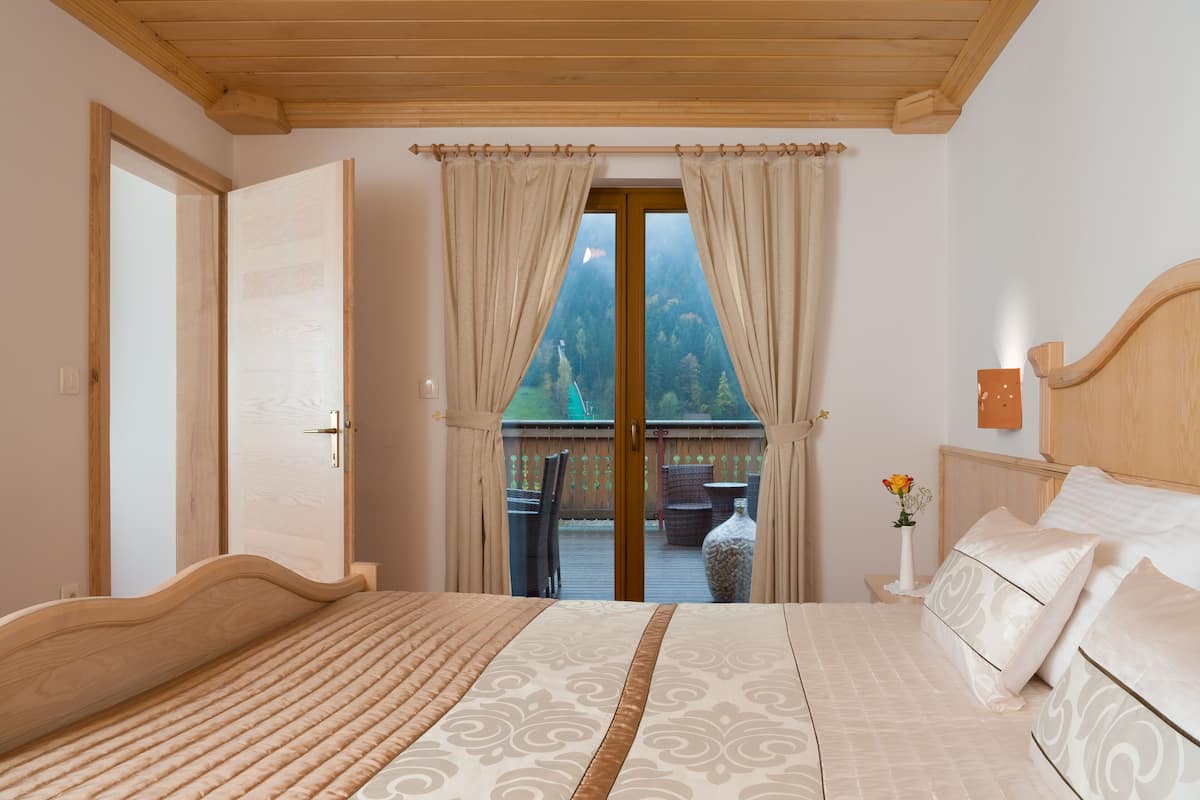 Quarto com cama grande, cabeceira de madeira e vista para uma varanda com mobiliário de exterior.