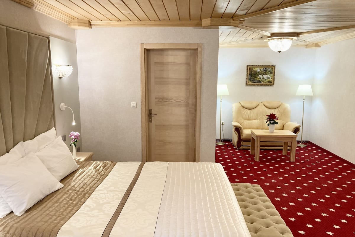 Quarto de hotel com uma cama grande, porta de madeira, tapete vermelho, sofá, duas luminárias e um quadro na parede.