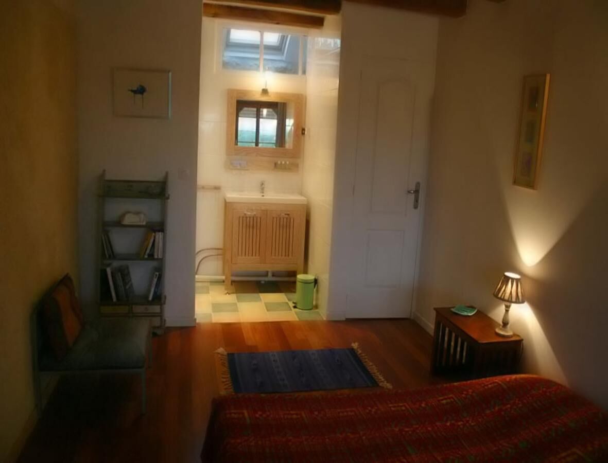 Um quarto aconchegante com piso de madeira, uma cama com manta vermelha, mesa de cabeceira com abajur, estante e janela com vista.