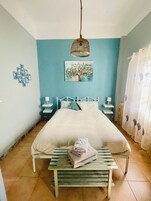 Quarto com cama, mesas de cabeceira, luminária pendente, pintura na parede e um banco com toalhas.