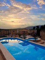 Piscina com espreguiçadeiras e guarda-sóis, cercada por área ajardinada com vista para colinas ao longe.