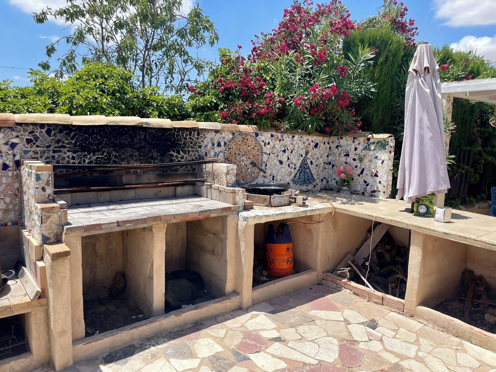 Uma área de cozinha externa com parede em mosaico, churrasqueira e um guarda-sol coberto.