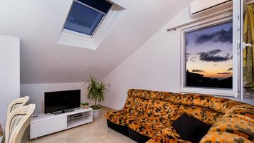Uma sala de estar moderna com televisão de tela plana, sofá estampado, planta em vaso e janela com vista para o pôr do sol.