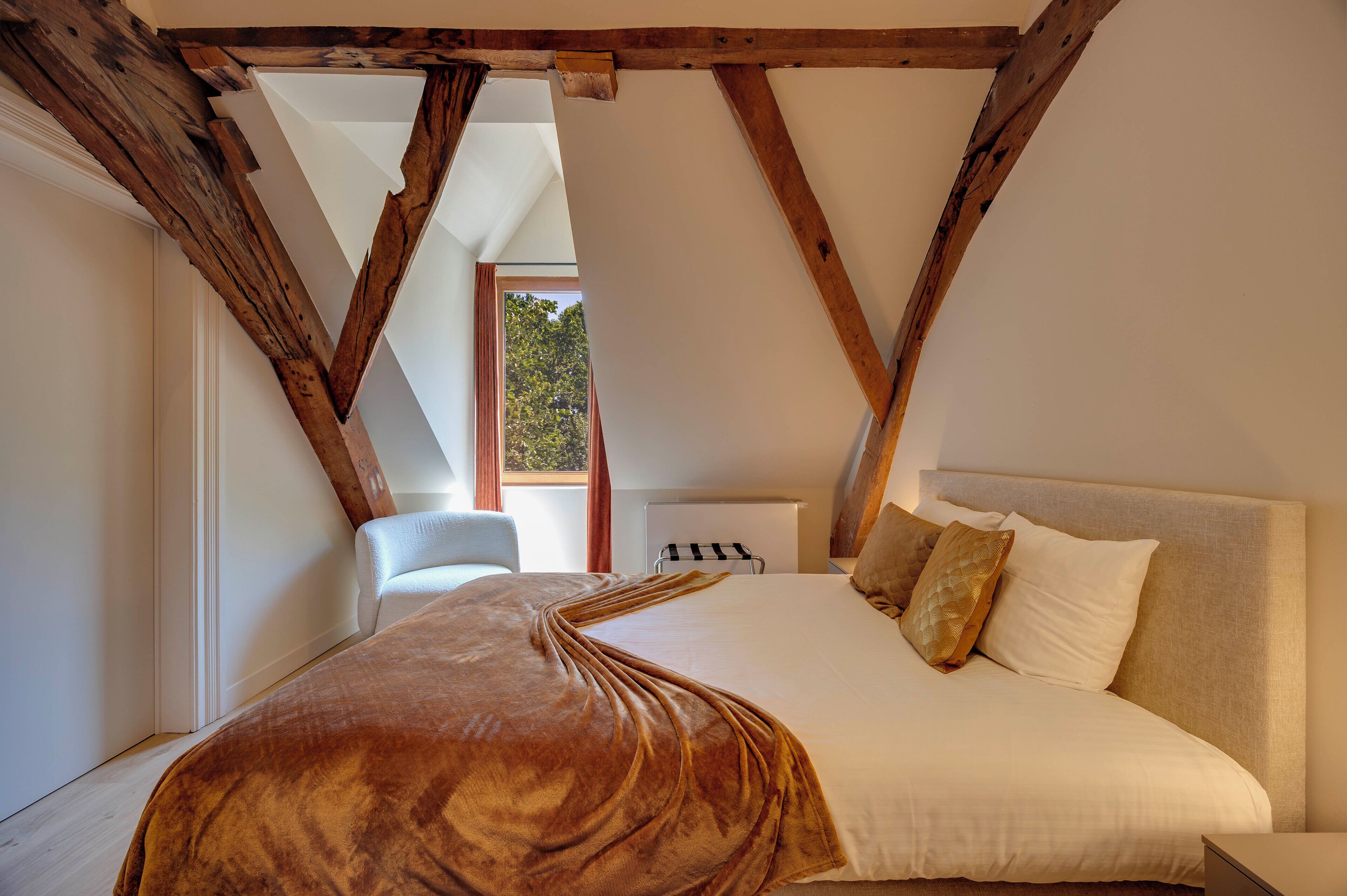 Quarto com cama grande, vigas de madeira, janela e vista para o verde lá fora.