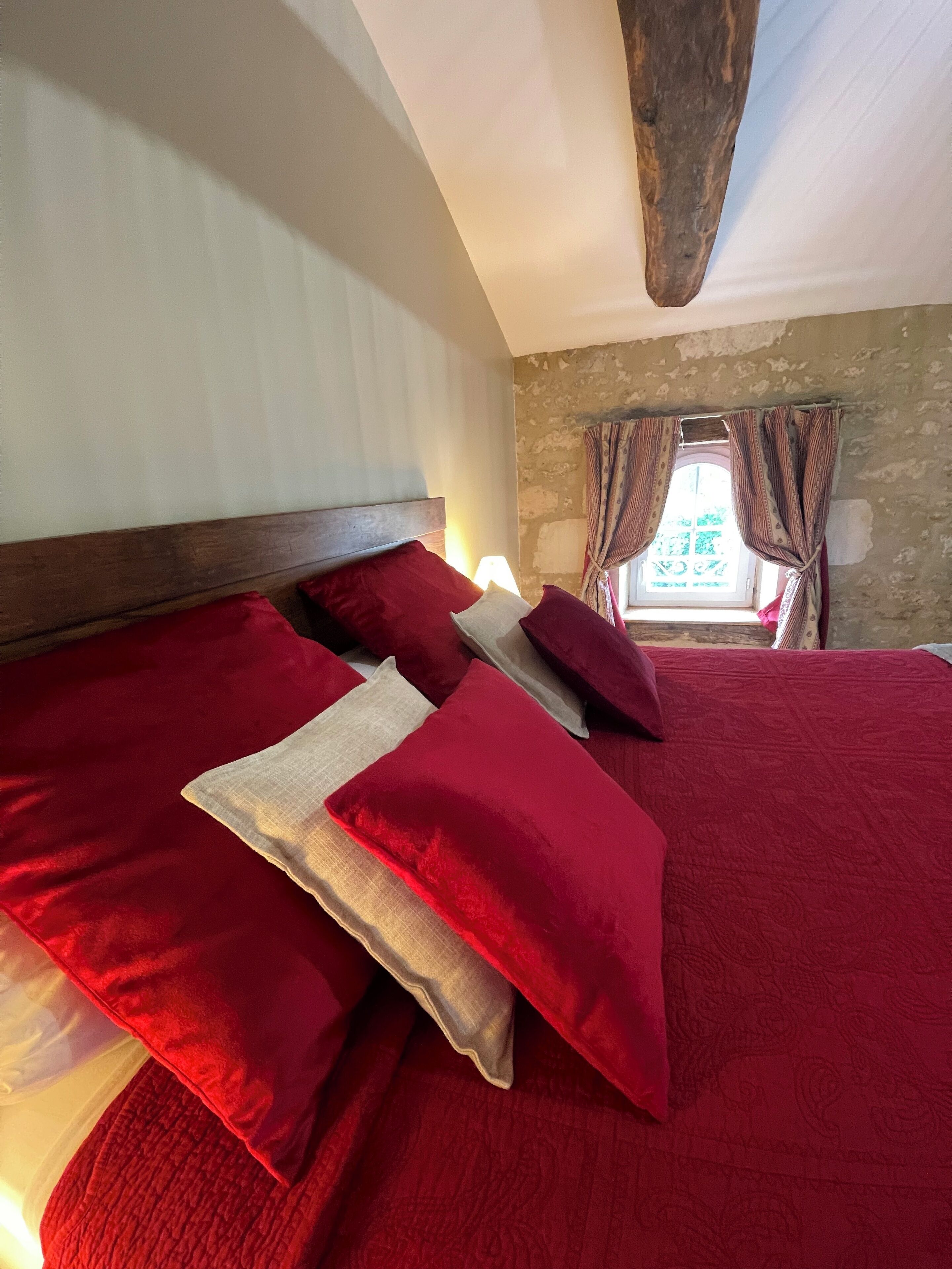 Um quarto com cabeceira de madeira, uma cama grande com roupa de cama vermelha e bege, uma mesa de cabeceira com abajur e uma janela com cortinas.