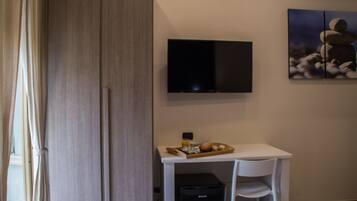 Quarto de hotel com televisão de tela plana na parede, uma pequena mesa com bandeja de petiscos, uma cadeira branca e uma lixeira.