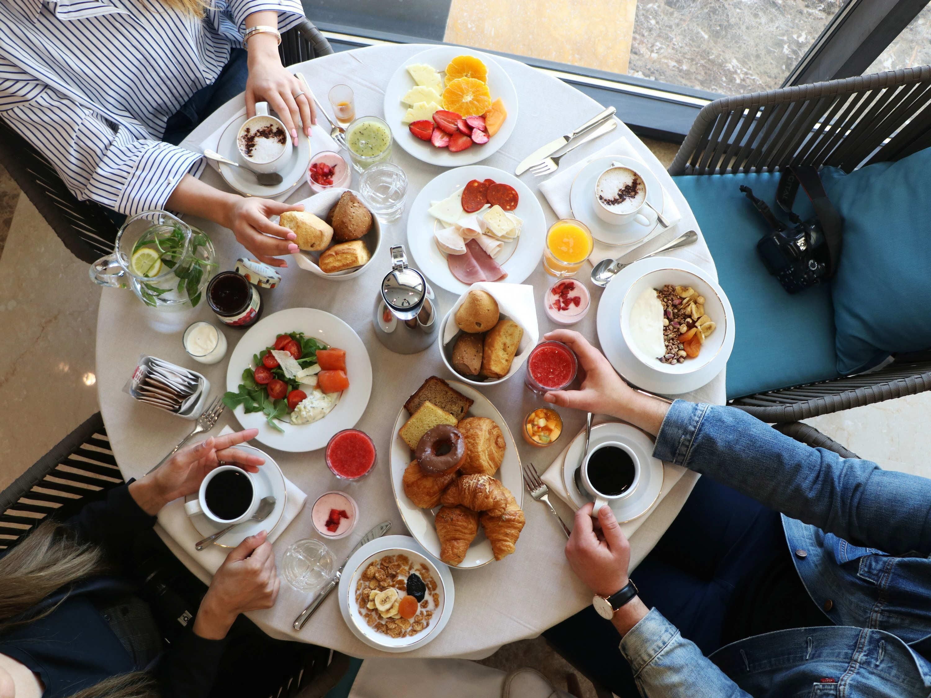 Mesa redonda com diversos itens de café da manhã, incluindo croissants, frutas e bebidas.