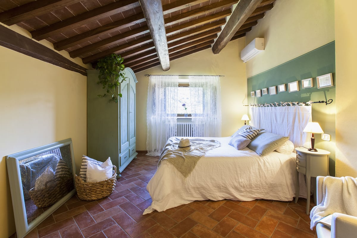 Quarto com cama, mesa de cabeceira com abajur, janela com cortinas, radiador e porta-retrato fixado na parede.