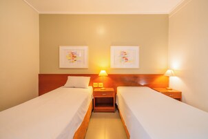 Quarto de hotel com duas camas de solteiro, cabeceiras de madeira, mesas de cabeceira com abajures e duas obras de arte emolduradas na parede.