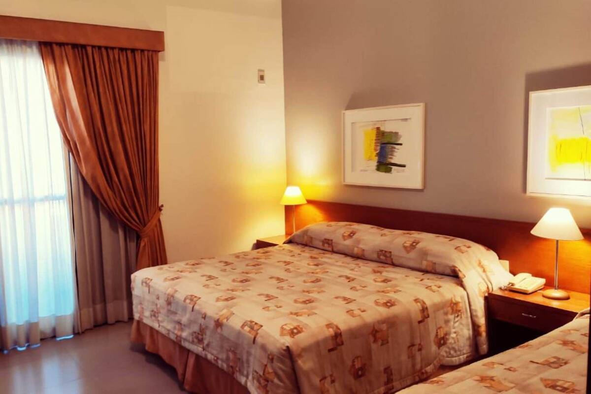 Quarto de hotel com cama, duas luminárias de cabeceira, uma janela com cortinas e um quadro na parede.