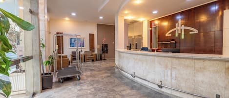 Uma área de recepção de hotel com balcão de mármore, mesa de atendimento e espaço de espera com cadeiras.