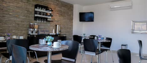 Uma zona de refeições moderna com cadeiras pretas, uma mesa redonda, uma parede de pedra, uma televisão na parede e um espaço para café.