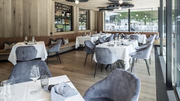 Um restaurante com várias mesas e cadeiras, paredes de madeira e amplas janelas que oferecem vista para o exterior.