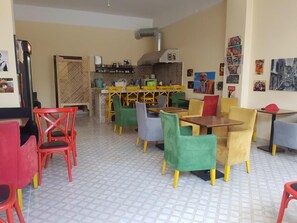 Uma área de refeições com cadeiras e mesas coloridas, uma cozinha ao fundo e diversas obras de arte nas paredes.