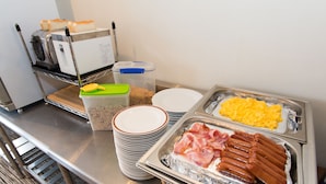 Buffet com diversos alimentos, incluindo cachorros-quentes, ovos mexidos e um recipiente com cereal.