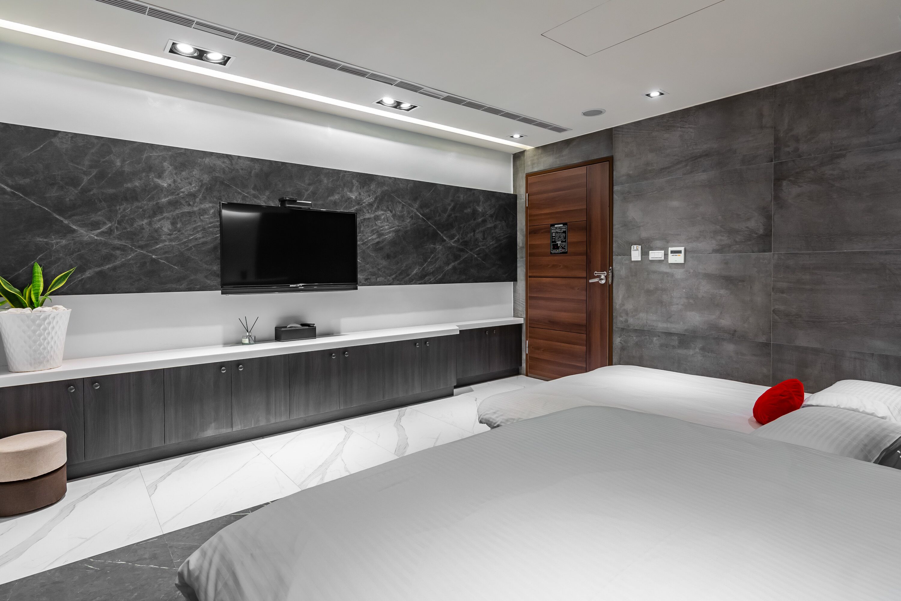 Quarto moderno com uma cama grande, uma televisão na parede, uma porta de madeira e um vaso com planta.