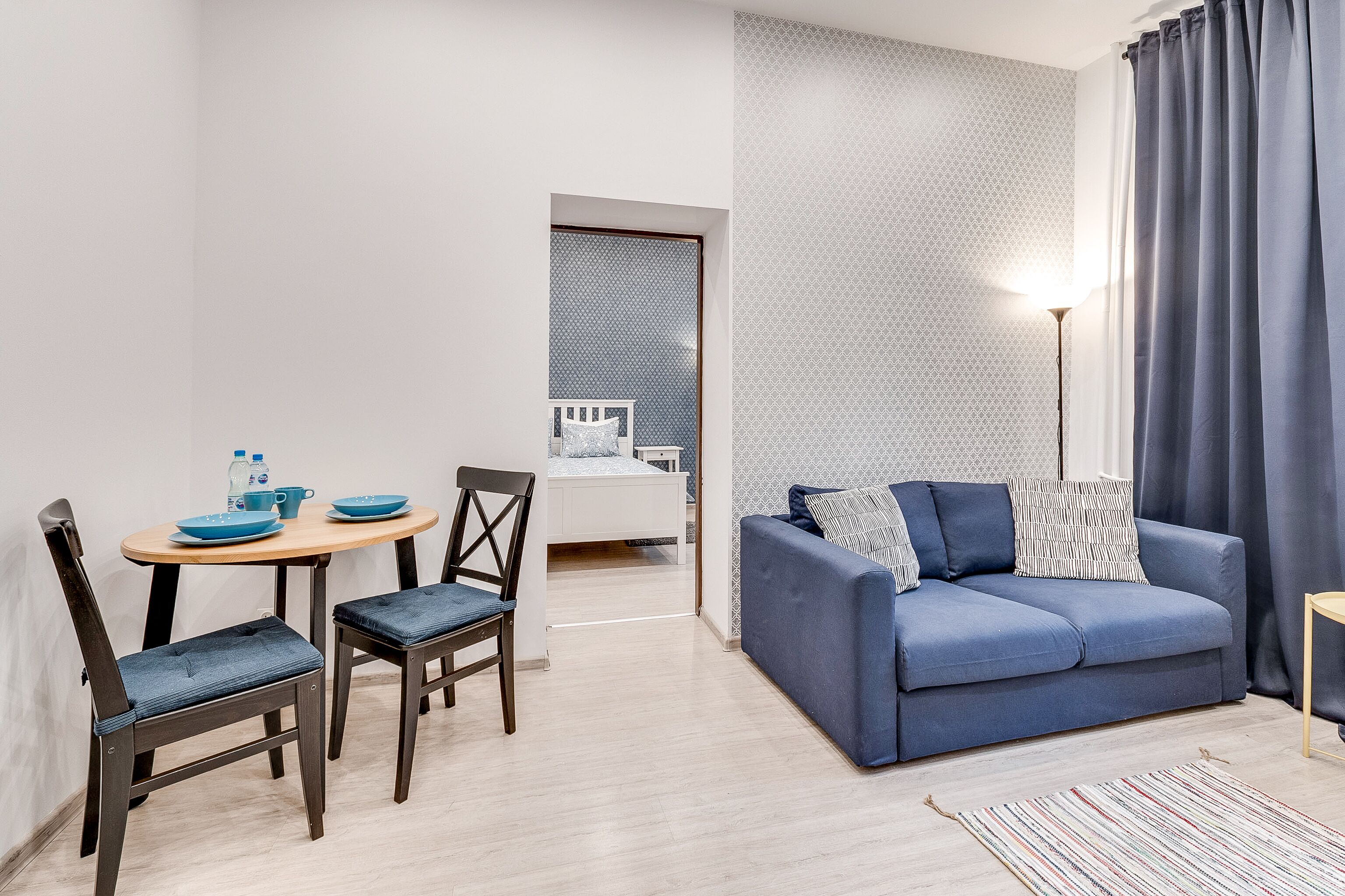 Uma sala de estar moderna com um sofá azul, duas cadeiras de madeira escura, uma mesinha redonda com pratos azuis e uma garrafa de água, e uma parede estampada.