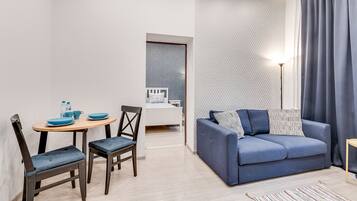 Uma sala de estar moderna com um sofá azul, duas cadeiras de madeira escura, uma mesinha redonda com pratos azuis e uma garrafa de água, e uma parede estampada.
