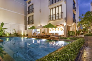 Área de piscina com espreguiçadeiras e guarda-sóis, cercada por vegetação e um edifício com varandas.