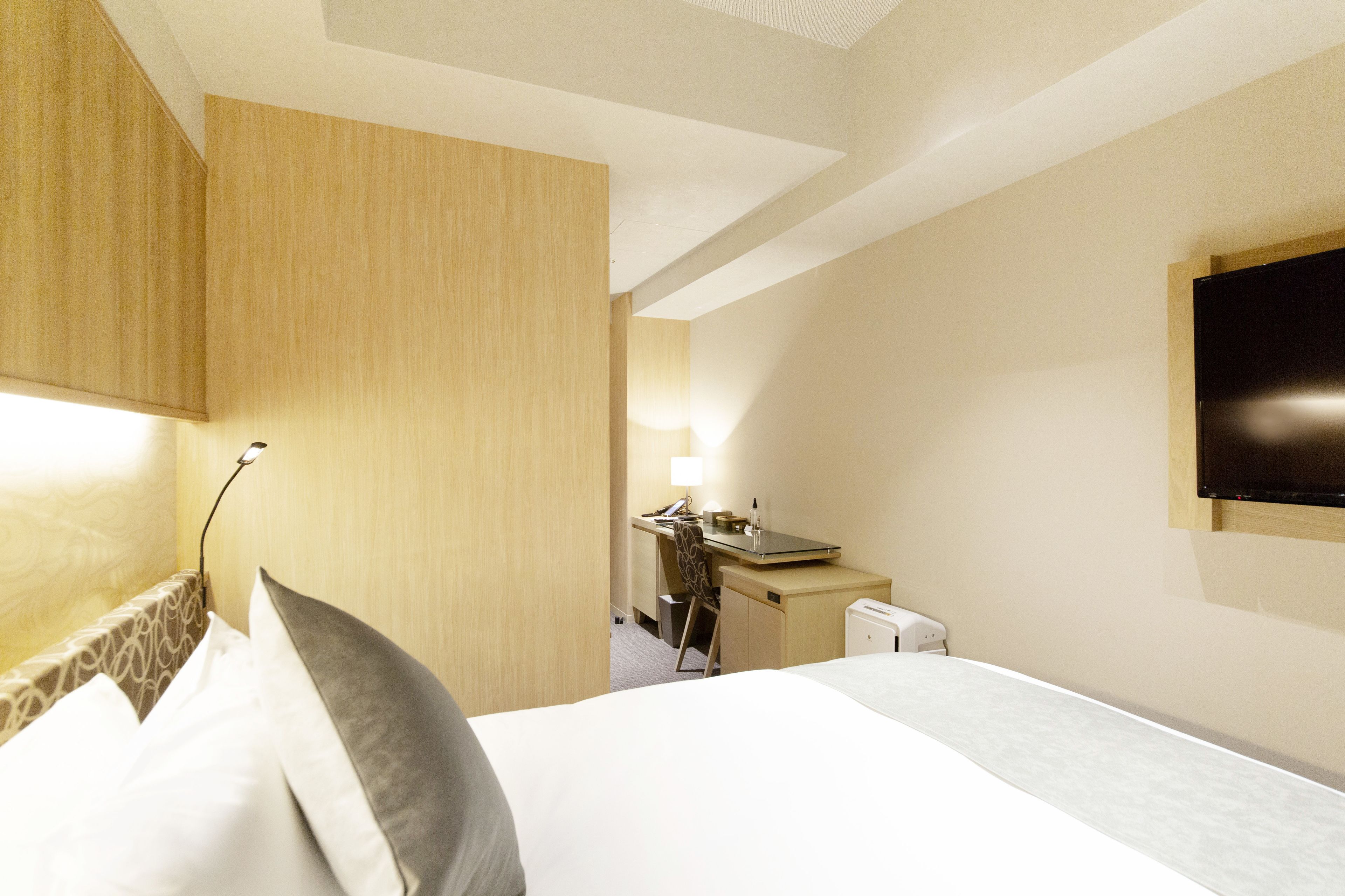 Quarto de hotel com cama, escrivaninha, cadeira, televisão e um eletrodoméstico branco pequeno.