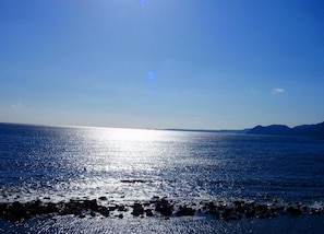 Visão clara de um mar calmo com costa rochosa e montanhas ao longe sob um céu azul brilhante.