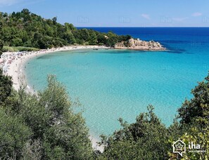 Uma praia costeira com águas turquesa cristalinas, areal arenoso e vegetação densa ao fundo.