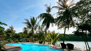 Piscina com deck de madeira, cercada por palmeiras e vista para um lago.