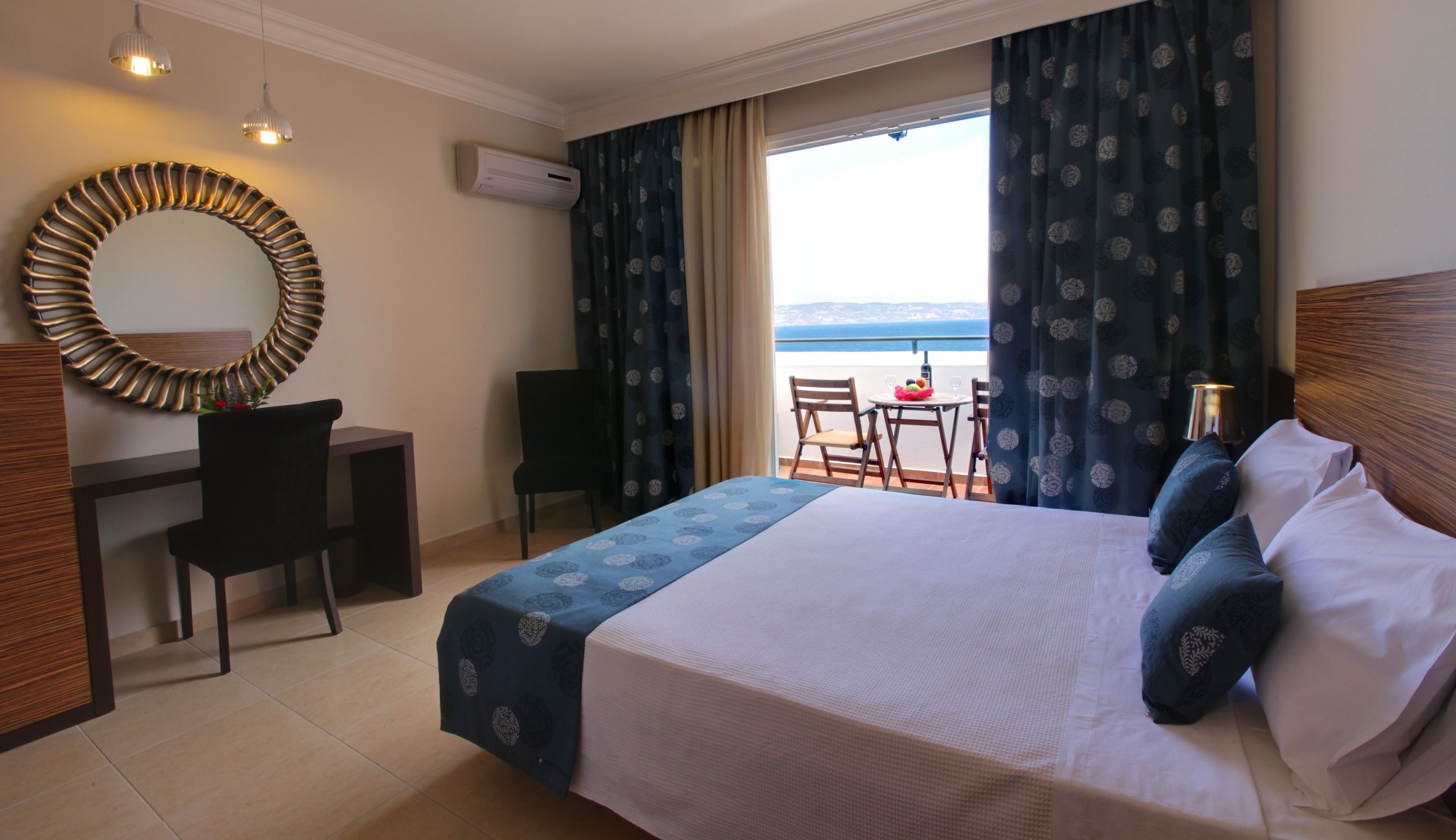 Quarto de hotel com cama, escrivaninha, cadeira, mesa com uma tigela de frutas e vista para o mar.