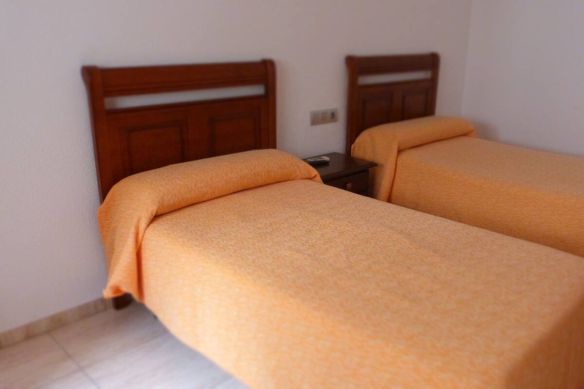 Duas camas de solteiro com cabeceiras de madeira e roupa de cama laranja num quarto pequeno com chão de azulejos.