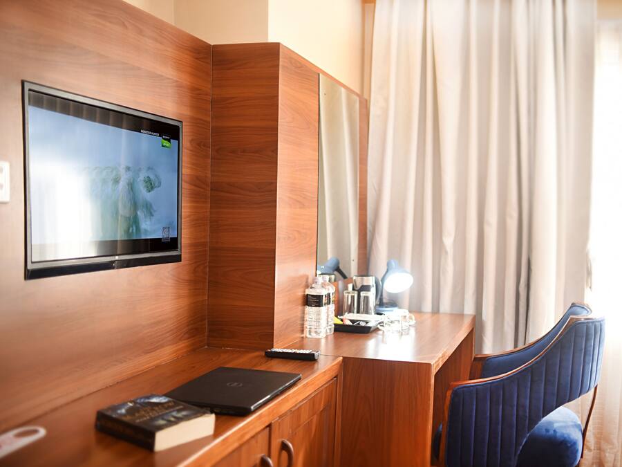 Quarto de hotel com secretária de madeira, televisão na parede, uma cadeira e janela com cortinas.
