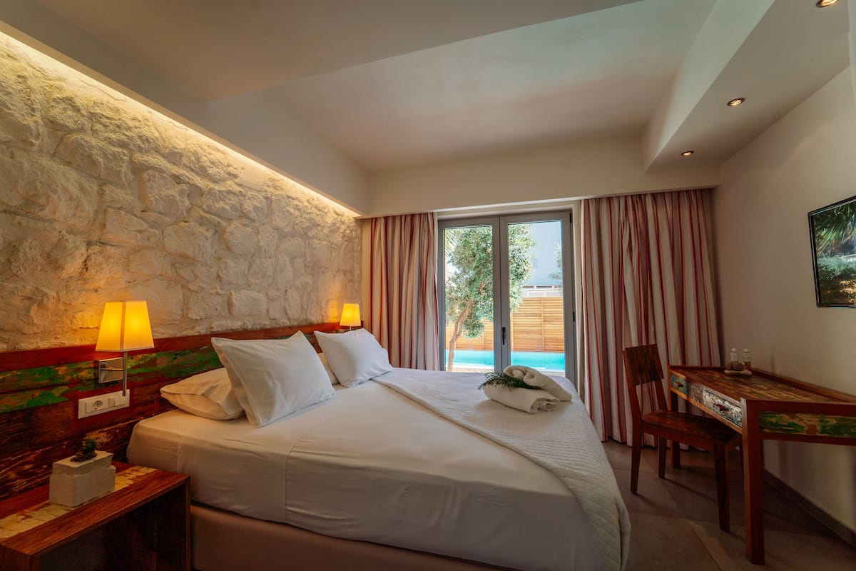 Quarto de hotel com parede de pedra, cama com roupa de cama branca, escrivaninha com cadeira e vista para o exterior.