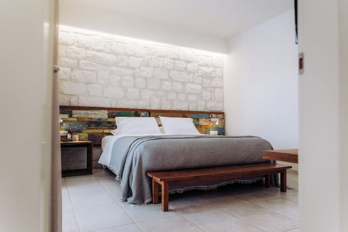 Quarto com cabeceira em parede de pedra, cama com roupa de cama branca, um banco de madeira e uma mesa de cabeceira com abajur.