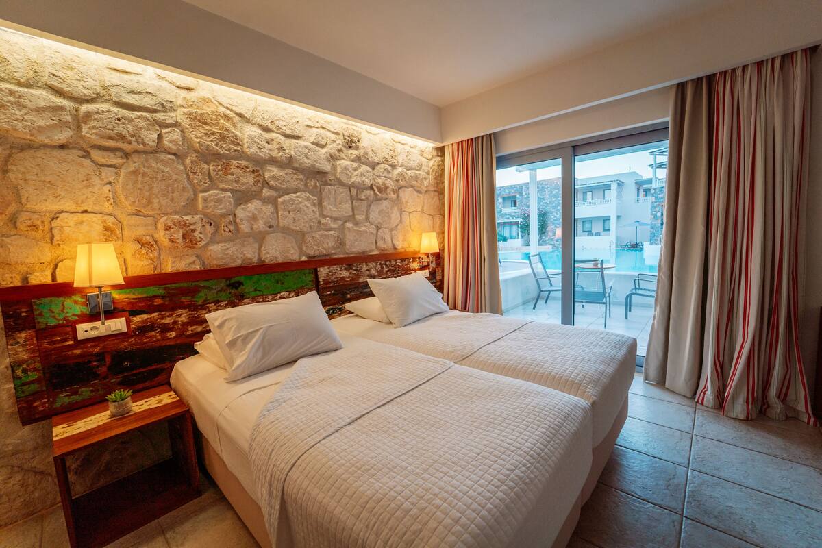 Quarto de hotel com parede de pedra, duas camas, uma mesa de cabeceira, abajur e vista para a varanda.