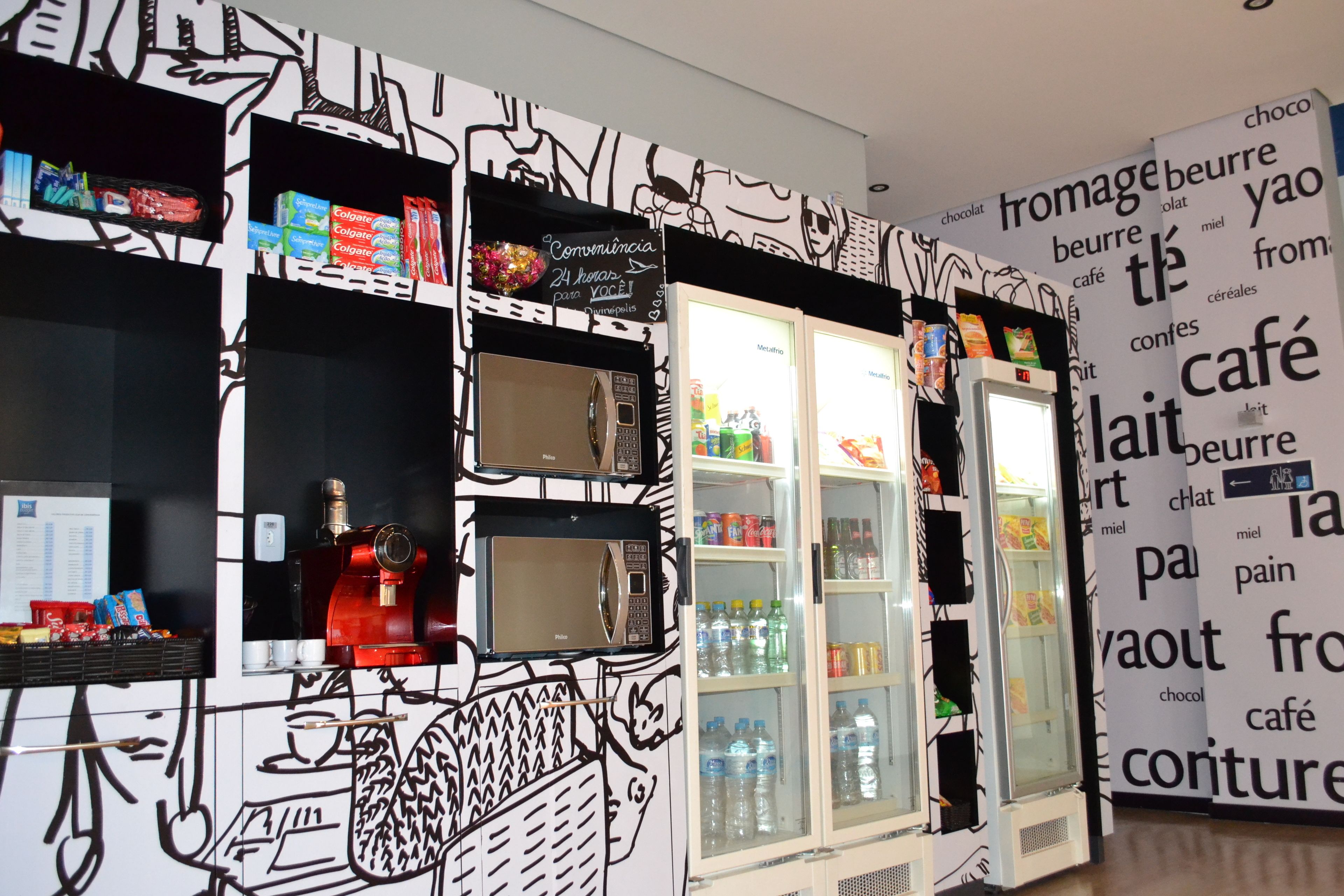 Um café com um mural em preto e branco, um refrigerador cheio de bebidas e prateleiras com lanches e temperos.