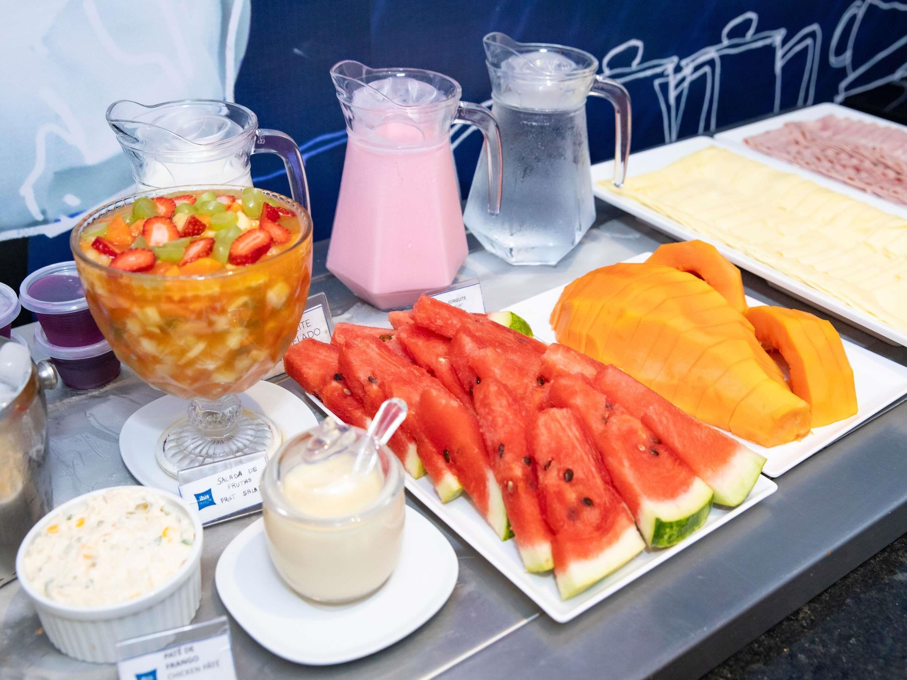 Uma mesa de buffet com diversos alimentos, incluindo salada de frutas, fatias de melancia e um jarro de água.