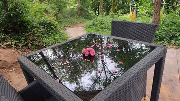 Mesa externa com tampo de vidro, rodeada por cadeiras de vime, em um jardim exuberante com um caminho de terra e vegetação ao fundo.