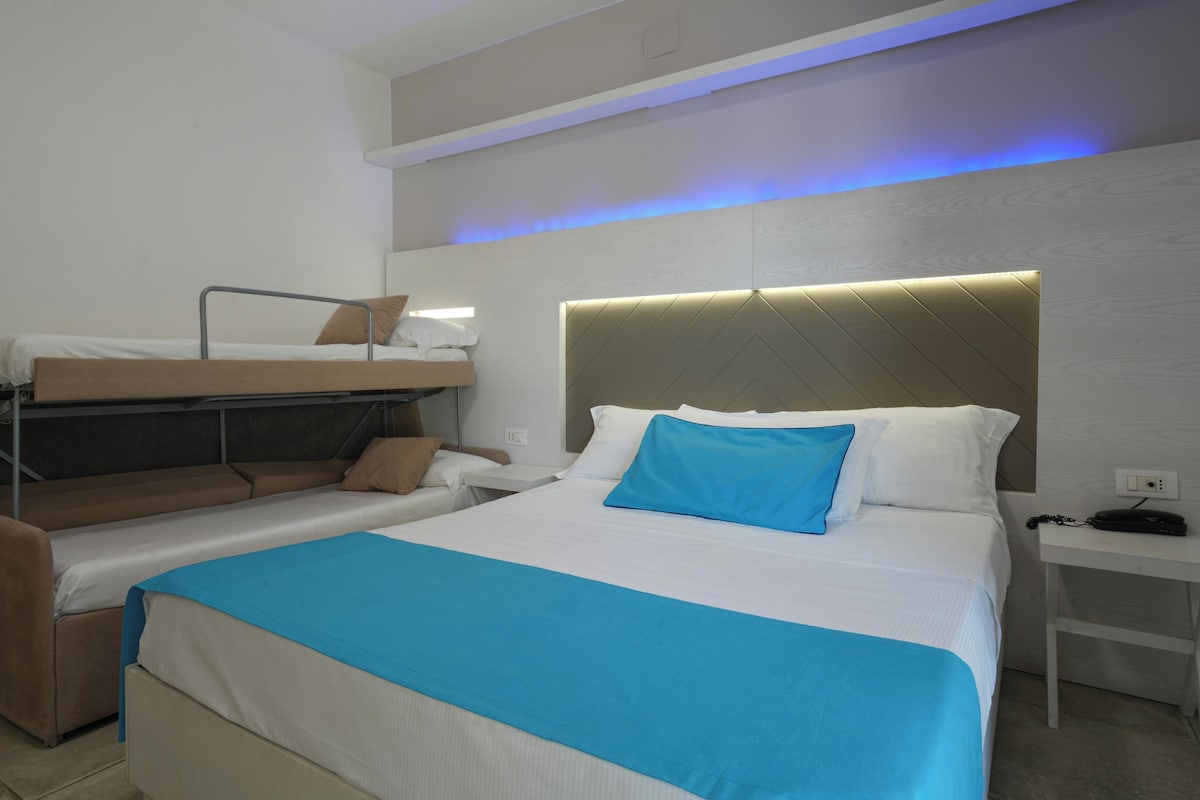 Quarto de hotel com beliche, uma cama de solteiro, mesa de cabeceira com telefone e luminária na parede.