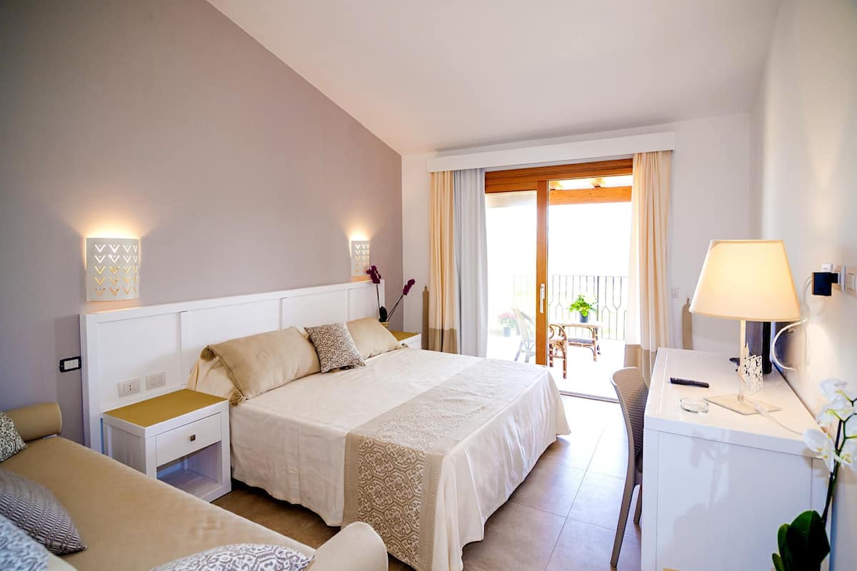 Quarto de hotel com uma cama grande, duas mesas de cabeceira, uma escrivaninha com uma luminária, uma cadeira e vista para a varanda externa.