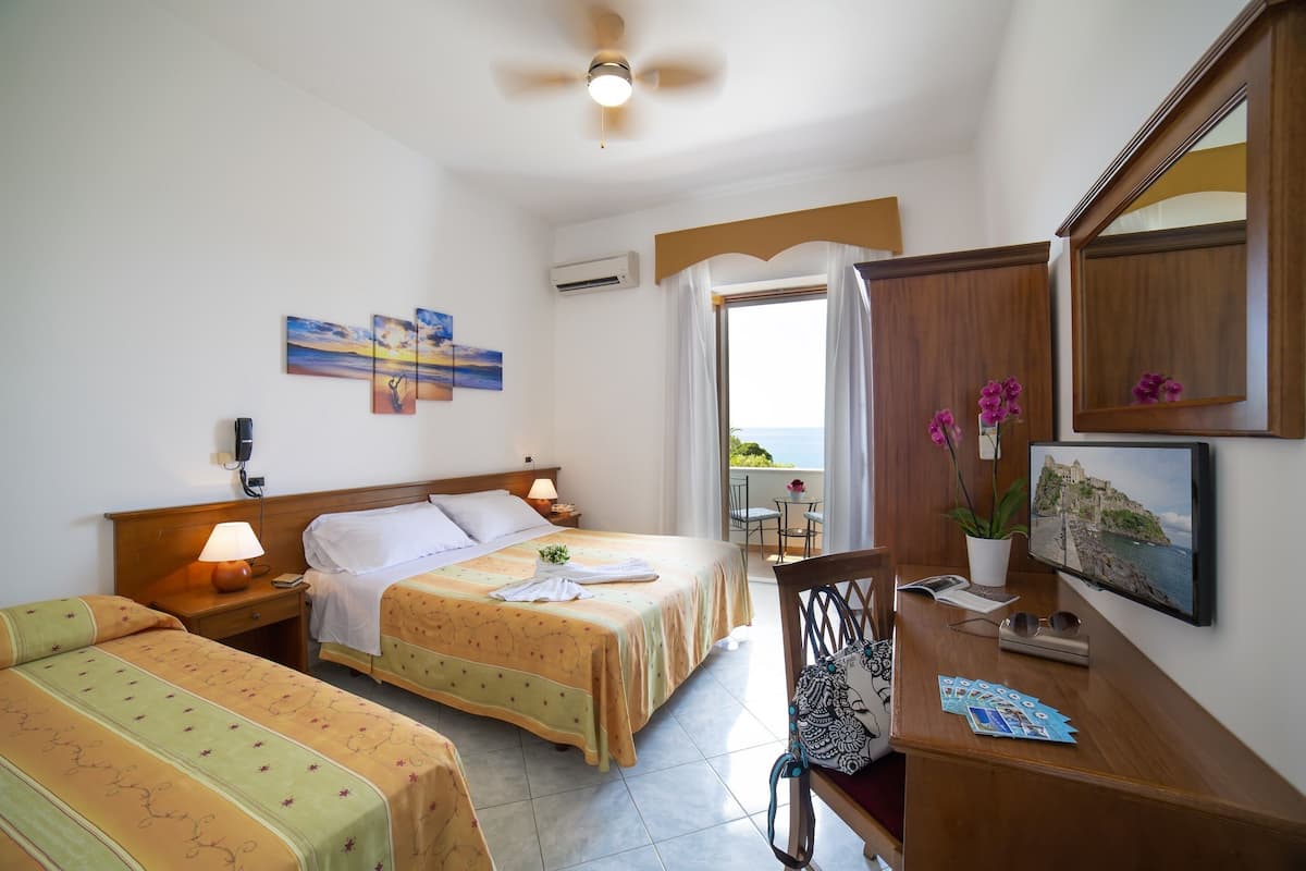 Quarto de hotel com duas camas, uma escrivaninha com cadeira, ventoinha no teto e varanda com vista.