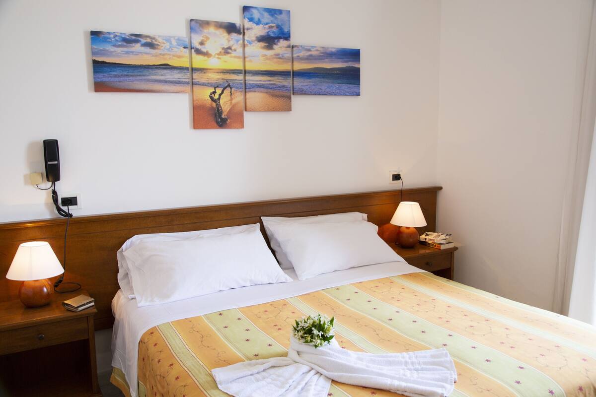 Quarto de hotel com cama, duas mesas de cabeceira, um criado-mudo, luminária, telefone e um quadro na parede.