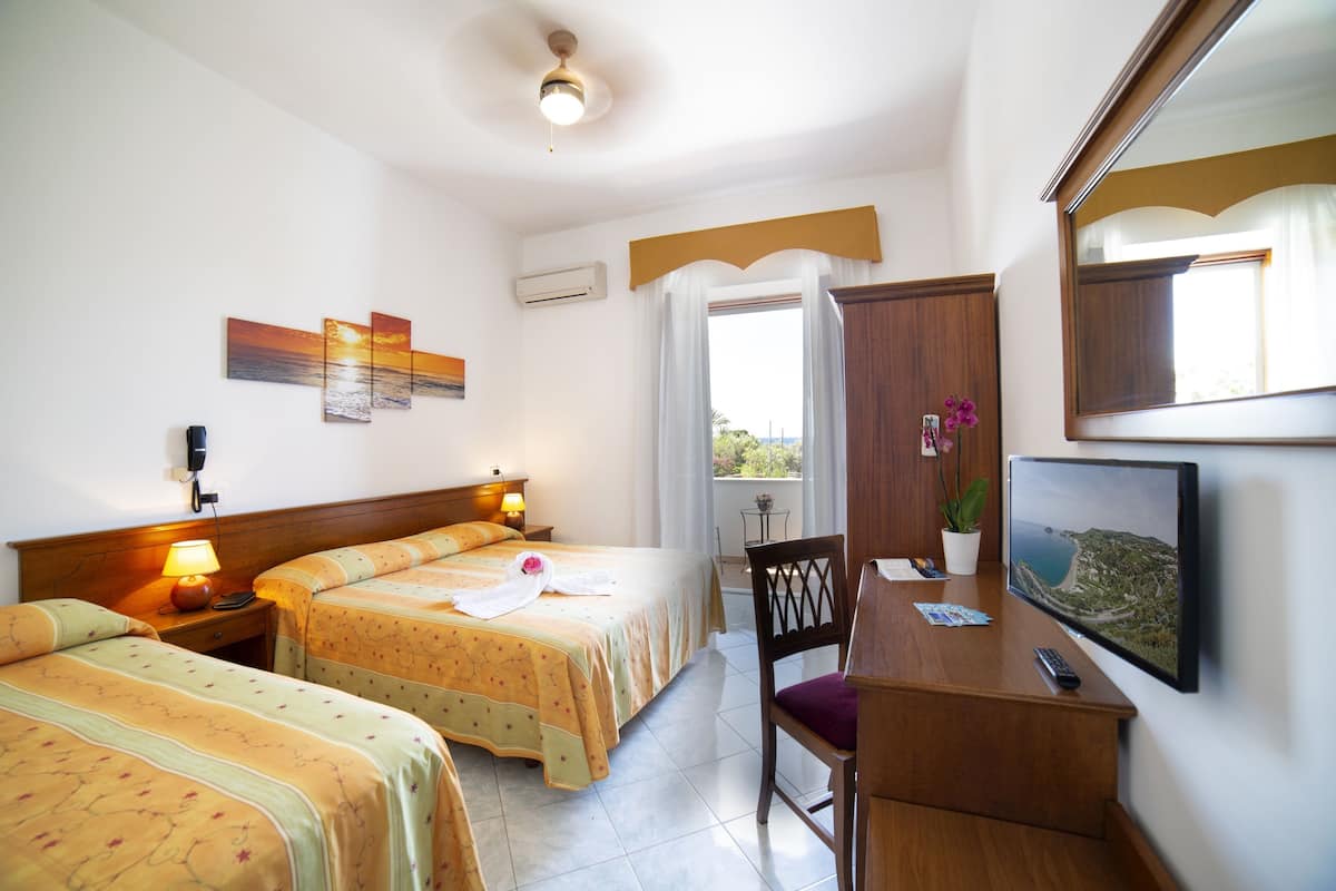Quarto de hotel com duas camas, uma mesa com televisão, uma cadeira, janela com cortinas e ventilador de teto.