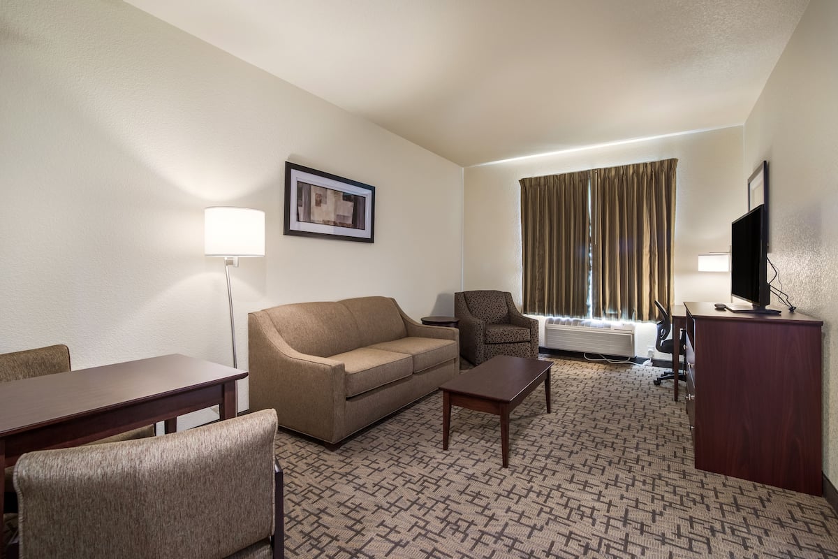 Quarto de hotel com sofá, poltrona, escrivaninha e televisão.