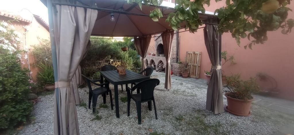 Pátio externo com mesa e cadeiras, cercado por plantas em vasos e uma parede de tijolos.