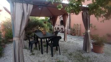 Pátio externo com mesa e cadeiras, cercado por plantas em vasos e uma parede de tijolos.
