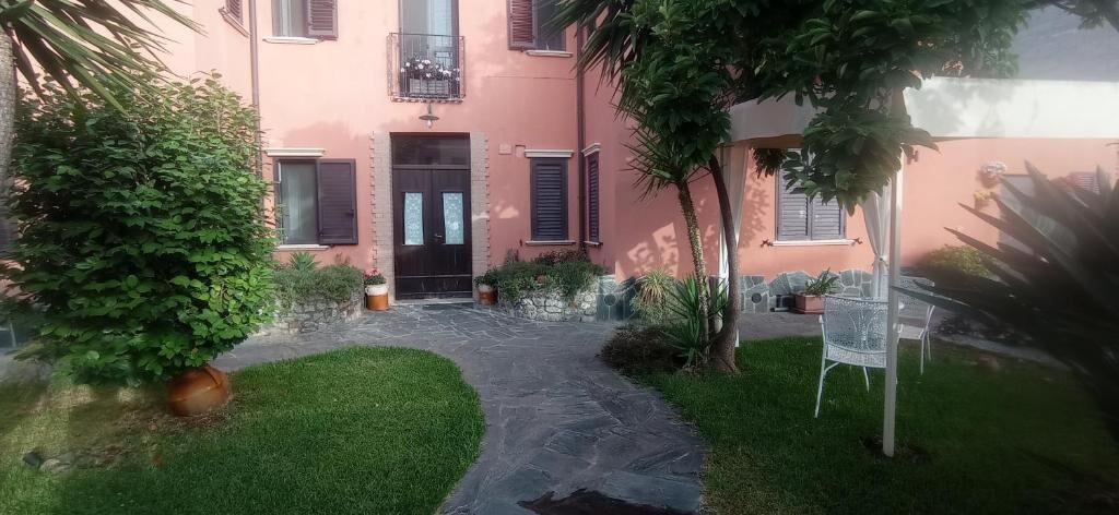 Um edifício rosa com porta preta, um caminho pavimentado e um pequeno jardim com plantas em vasos.